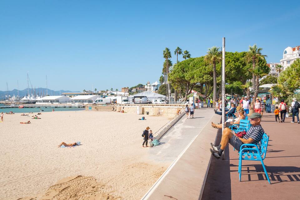 Uma praia com pessoas relaxando, um calçadão com bancos e um porto com barcos.