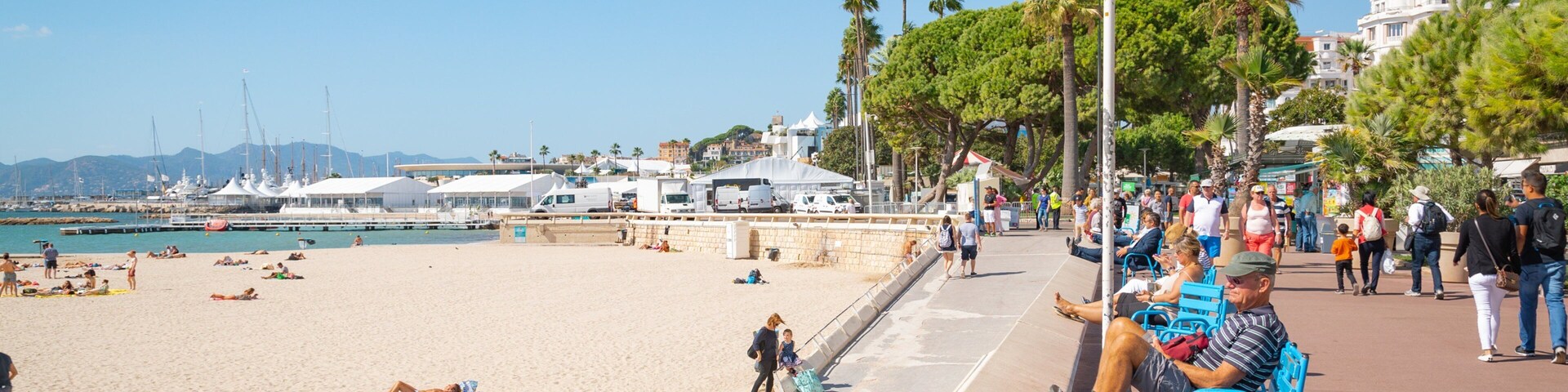 Uma praia com pessoas relaxando, um calçadão com bancos e um porto com barcos.