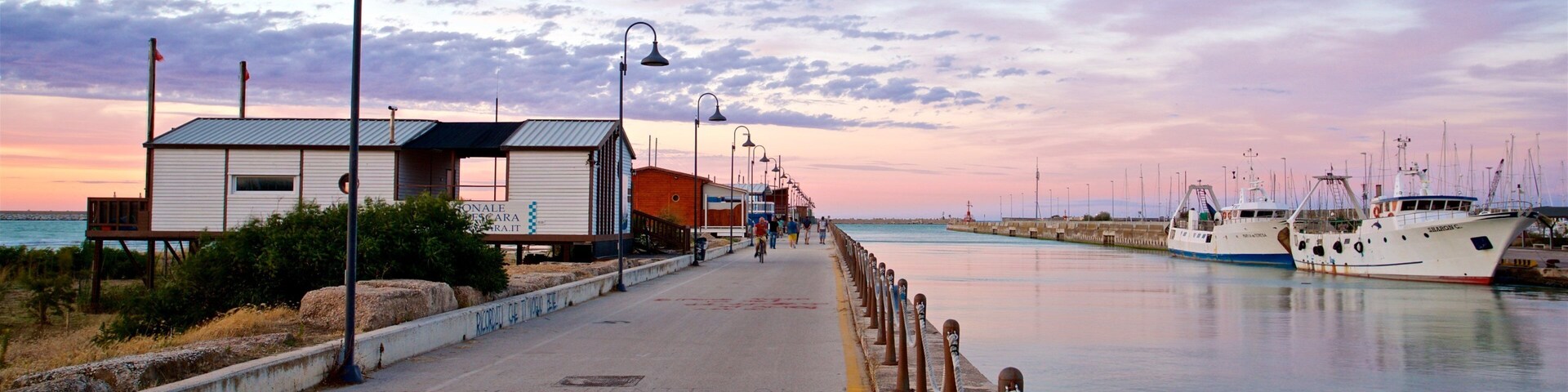 Um calçadão à beira-mar com uma fileira de edifícios, mar calmo e barcos atracados no cais.
