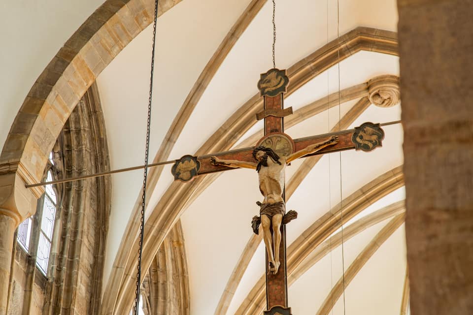 Cena da crucificação em uma catedral, com altos tetos abobadados e colunas de pedra.