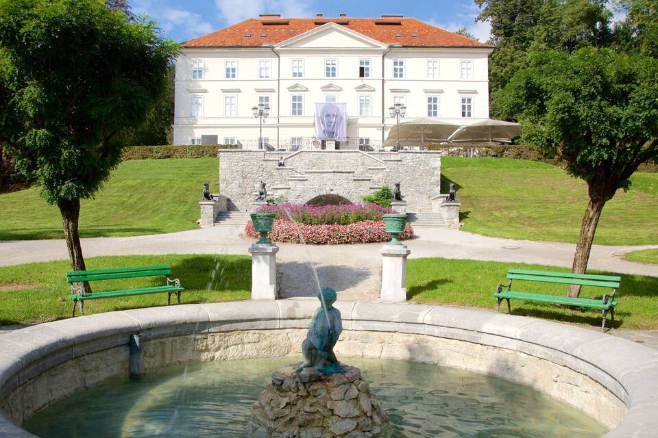 Uma fonte com uma estátua no centro, cercada por um muro de pedra. À frente da fonte, há uma estátua de um homem. Atrás da fonte, ergue-se um edifício grande e branco com telhado vermelho.
