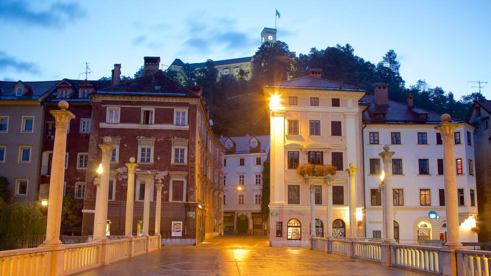Uma praça histórica na cidade com arquitetura clássica, uma torre do relógio imponente e montanhas ao fundo.