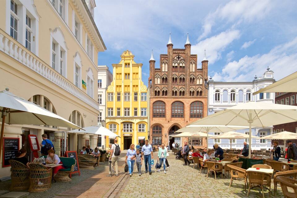Uma praça de calçamento com cafés ao ar livre, edifícios históricos e pessoas caminhando.