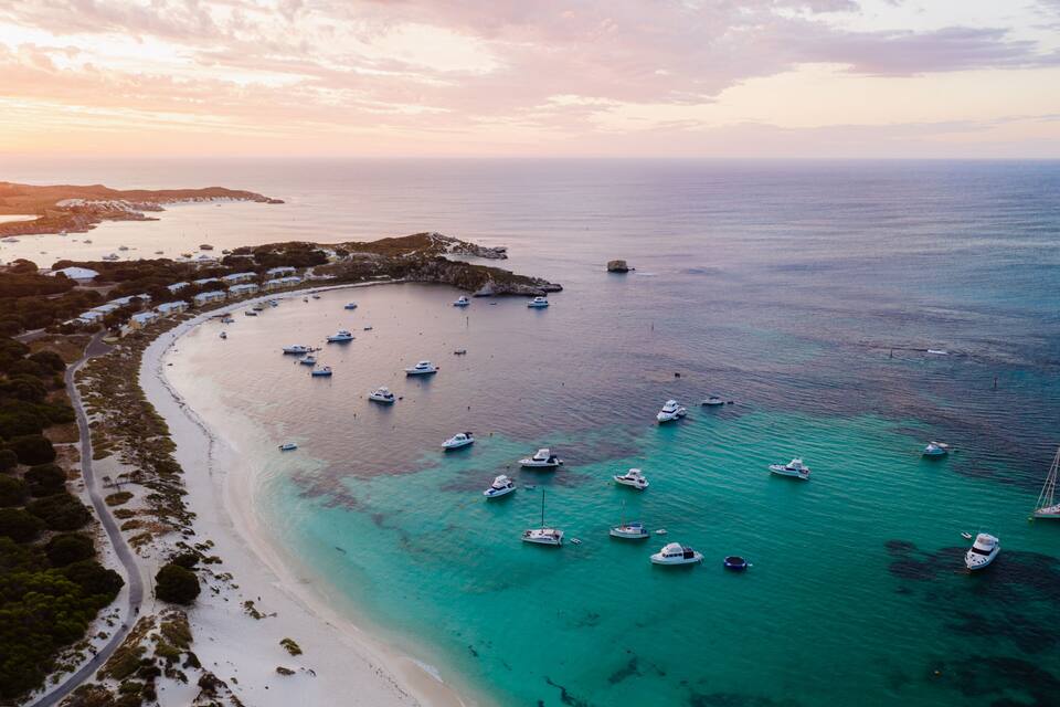 Uma área costeira com praia de areia, barcos ancorados na água e um afloramento rochoso ao longe.