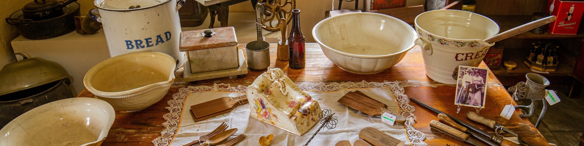 Uma cozinha antiga com utensílios de cozinha antigos, incluindo um recipiente para pão, uma tigela grande e vários utensílios sobre uma mesa com renda.