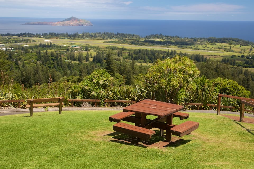 Mesa de piquenique com bancos numa área gramada, com vista panorâmica da paisagem costeira.