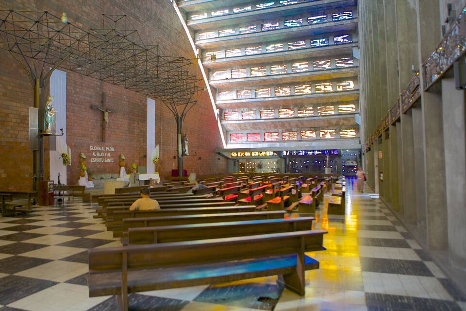 Interior de igreja com bancos de madeira, piso xadrez, parede de tijolos e uma grande janela de vitrais.