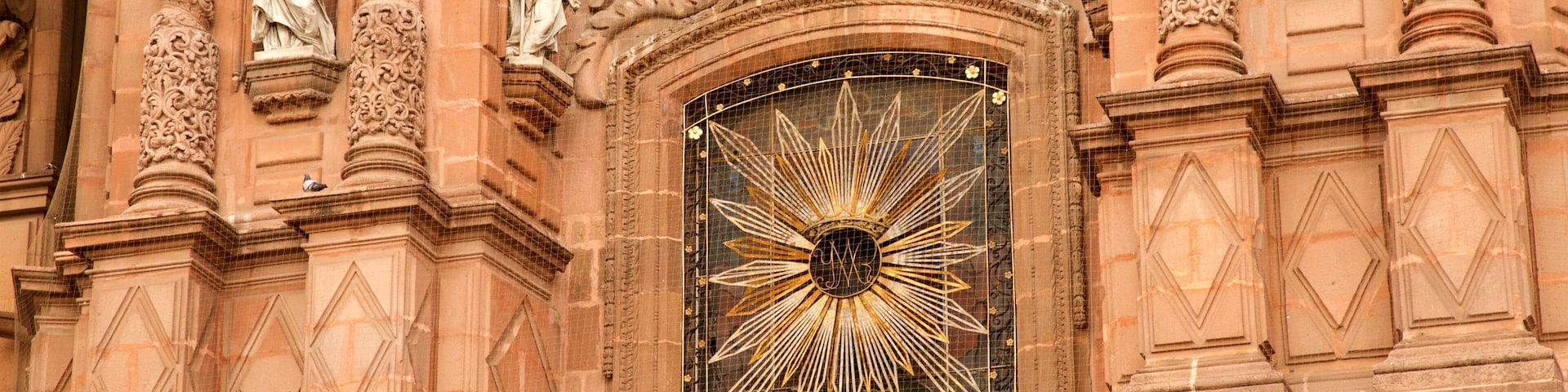 Fachada detalhada de um edifício histórico com esculturas elaboradas, estátuas e uma grande janela de vitrais com um desenho em forma de sol.