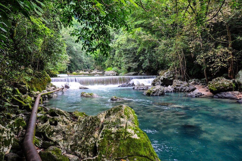 Uma cascata numa área arborizada, com águas cristalinas e arredores rochosos.
