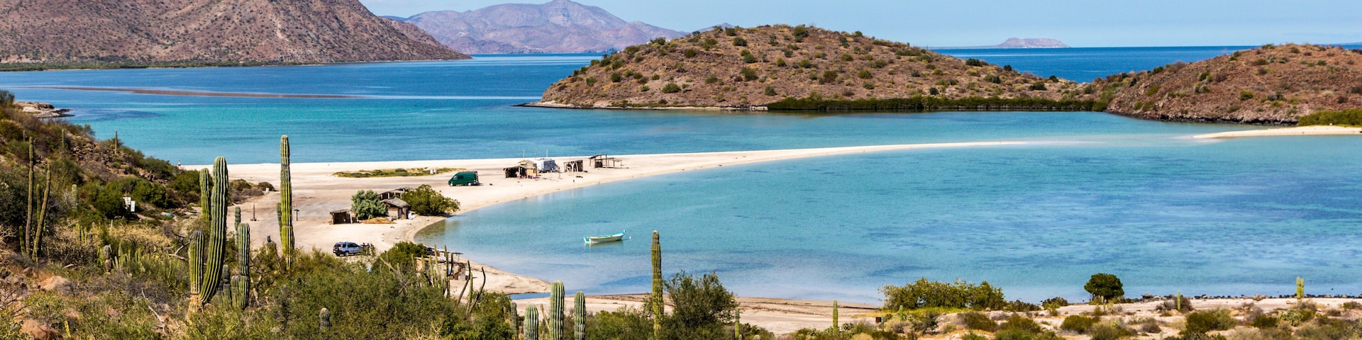 Paisagem costeira com uma praia arenosa, cactos e uma montanha ao fundo.