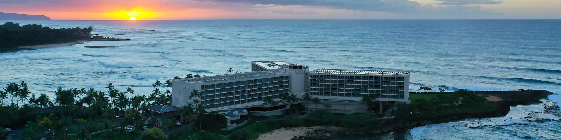 Um resort costeiro com hotel na orla, palmeiras e um pôr do sol sobre o oceano.