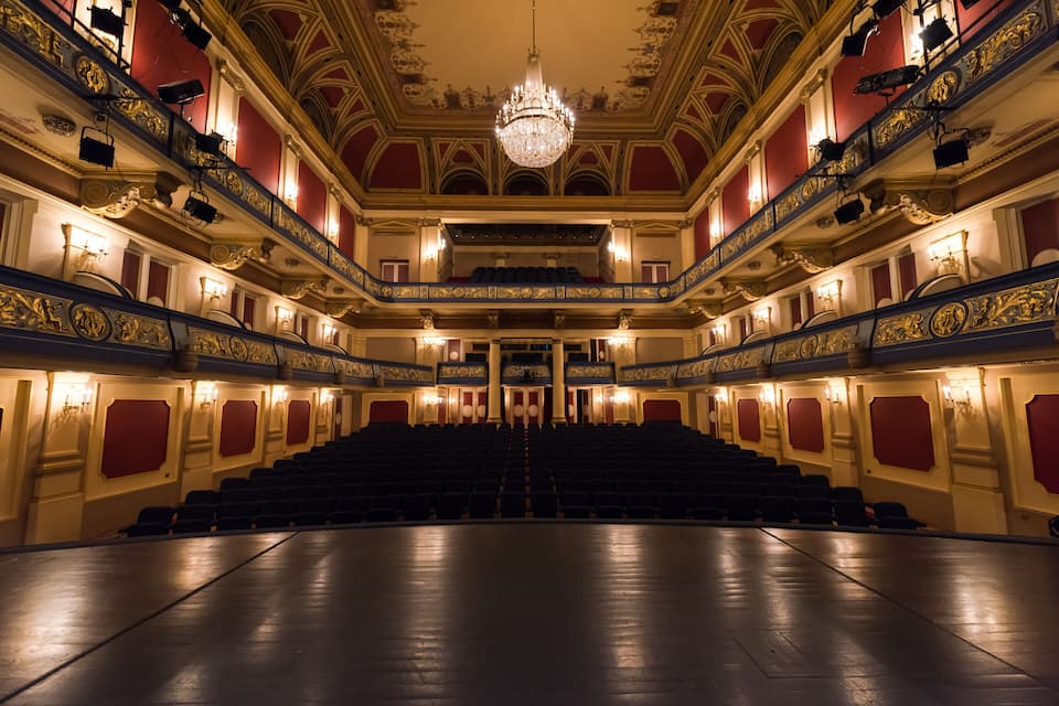 Um teatro ornamentado com um lustre imponente, várias galerias e fileiras de assentos vazios.