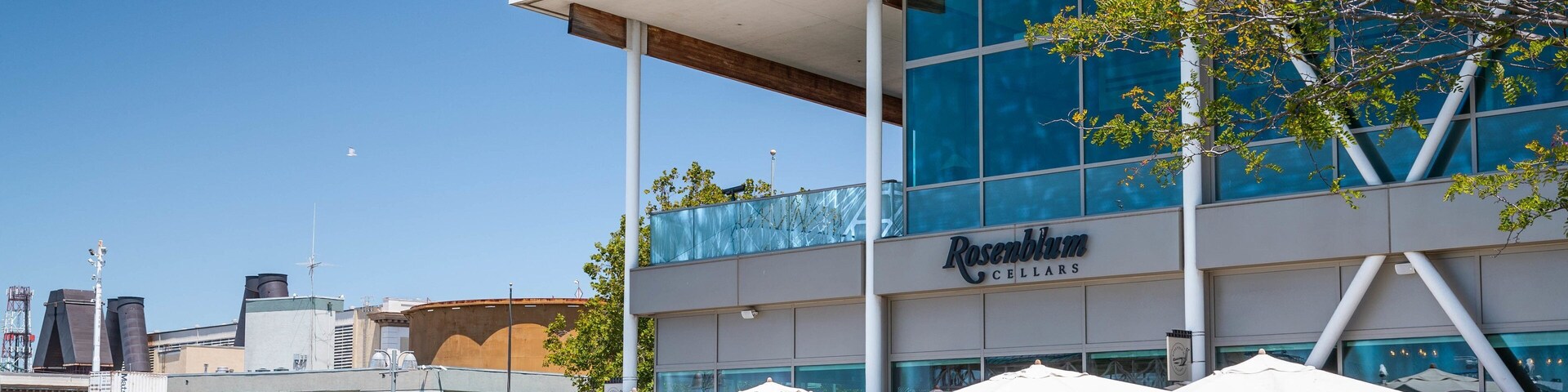 Um edifício moderno com grandes janelas e uma placa que diz "Rosenblum Cellars". Há guarda-sóis e vasos com flores em frente ao edifício.
