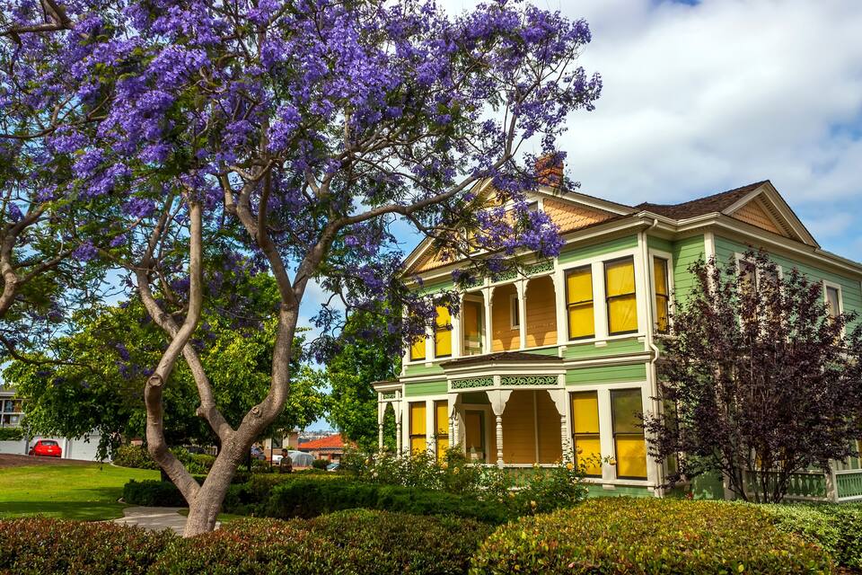Uma casa de dois andares com detalhes em verde e amarelo, cercada por jardins bem cuidados e uma grande árvore com flores roxas.