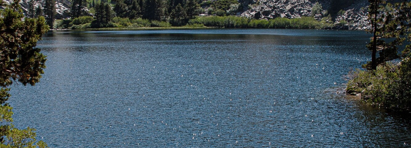 Um grupo de pessoas caminhando em um lago, rodeado por montanhas rochosas e árvores.