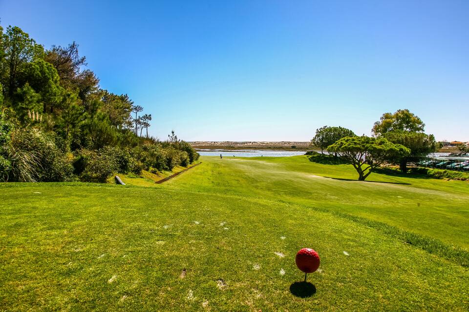Um campo de golfe com uma bola vermelha no green, rodeado por árvores e um céu azul límpido.