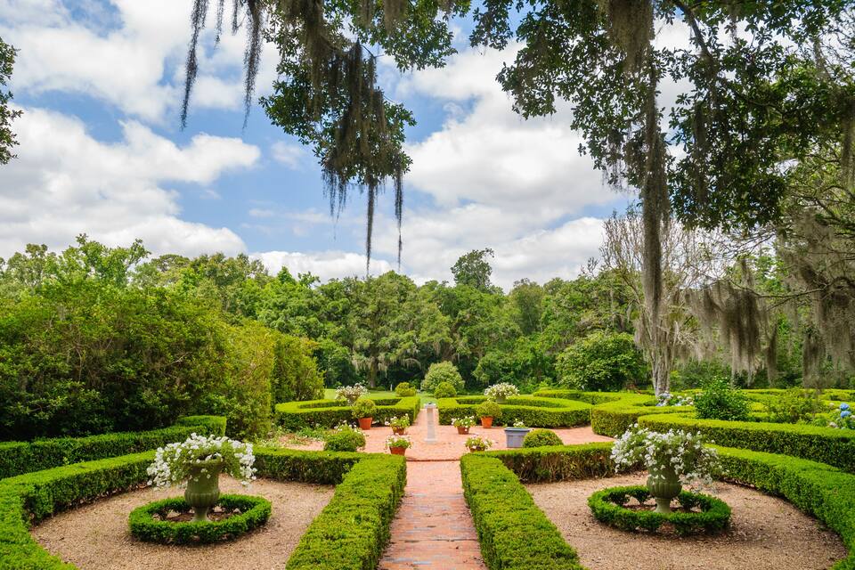 Um jardim formal com um caminho central, rodeado por sebes bem aparadas e plantas floridas.