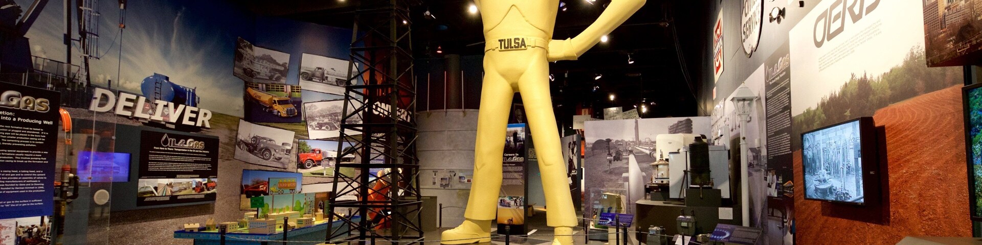 Uma grande estátua amarela de um homem de terno, com a inscrição 'TULSA' nas costas, cercada por painéis informativos e exposições.