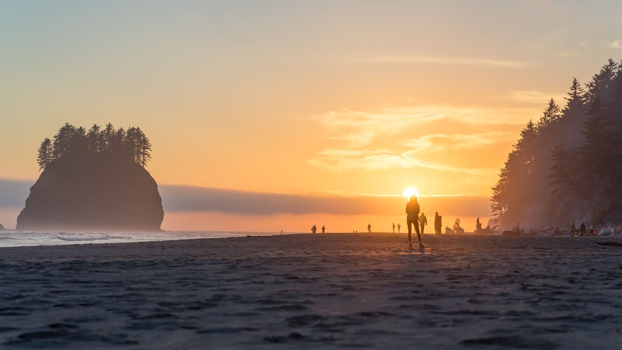 Uma praia ao pôr do sol, com uma grande formação rochosa e árvores.