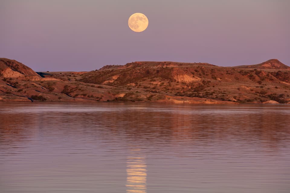 Paisagem iluminada pela lua, com um corpo d'água calmo refletindo a lua e as colinas ao redor.