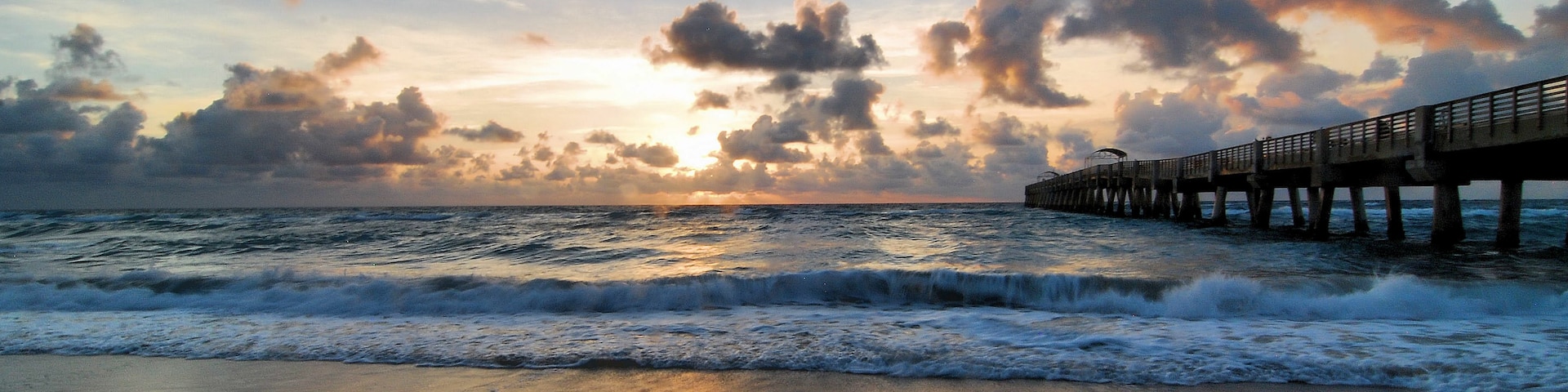 Uma praia com ondas ao pôr do sol, um píer que se estende para a água e um céu nublado.