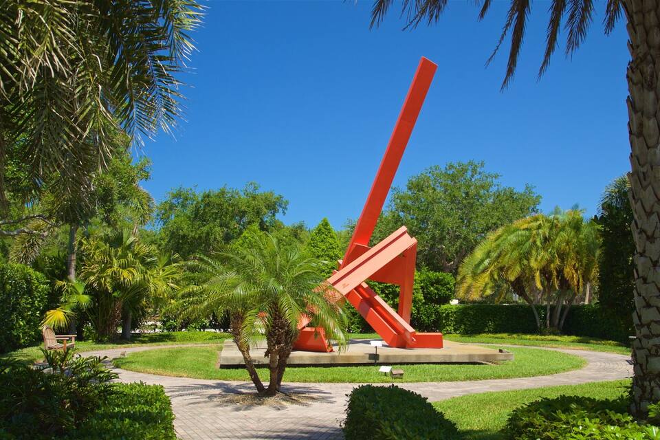 Escultura abstrata vermelha em um parque com palmeiras e um céu azul claro.