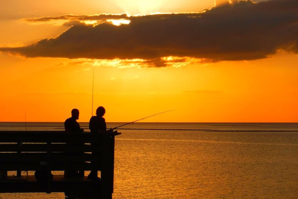 Um pôr do sol sobre a água, com duas pessoas pescando num cais.
