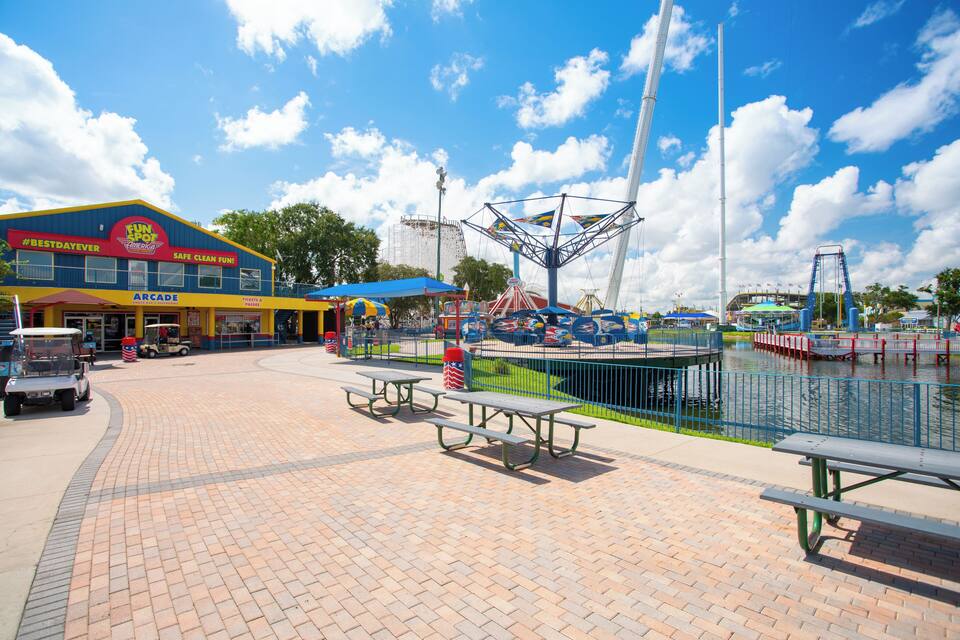 Um parque aquático com mesas de piquenique, uma roda gigante e um edifício com a inscrição 'Fun Spot Arcade'.