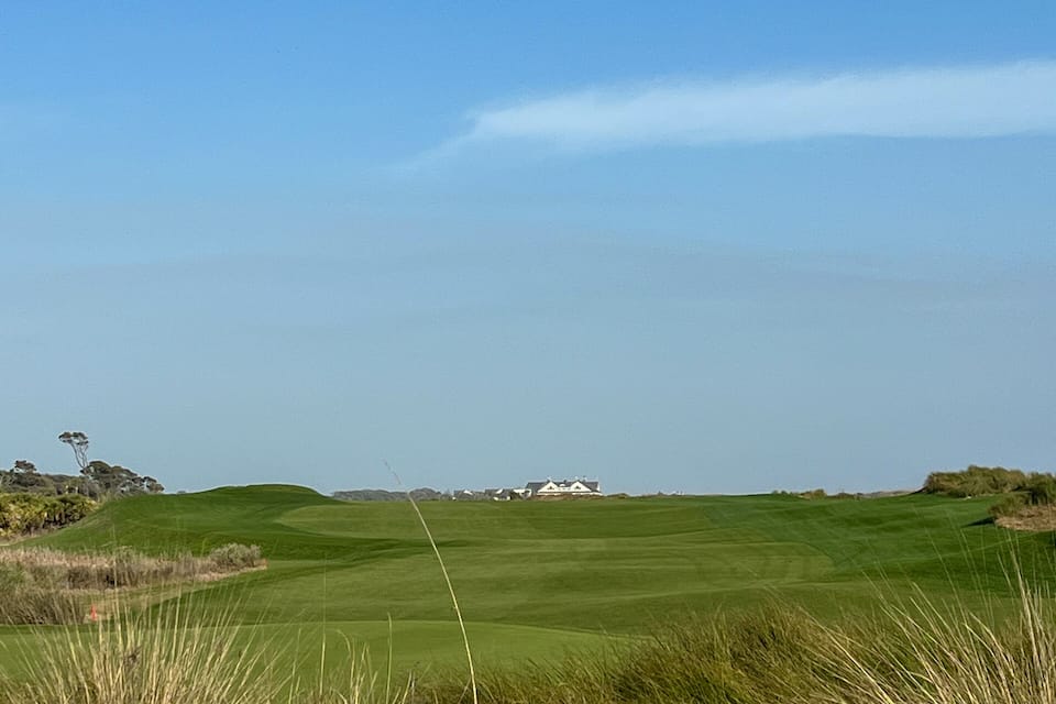 Um campo de golfe com céu azul límpido, grama verde e alguns edifícios ao longe.