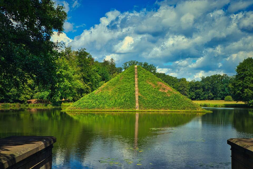 Uma ilha em formato de pirâmide em um lago tranquilo, rodeada por vegetação exuberante e um céu parcialmente nublado.