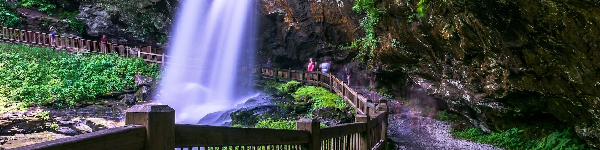 Uma cascata desce por um penhasco rochoso, rodeada por vegetação exuberante. Uma passarela de madeira com guarda-corpo acompanha o curso da água.
