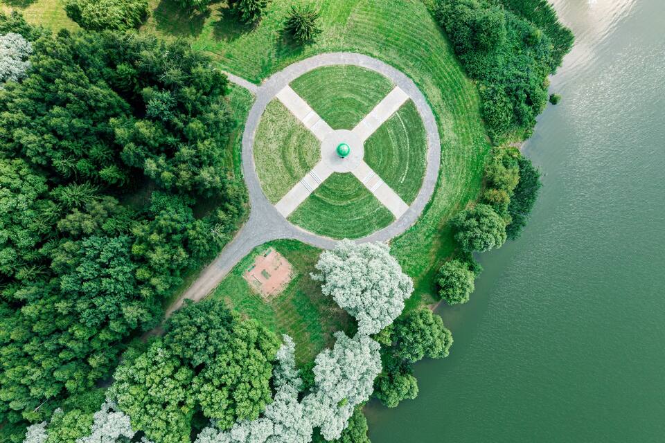 Vista aérea de um parque com um espaço verde circular, um lago e árvores densas ao redor.