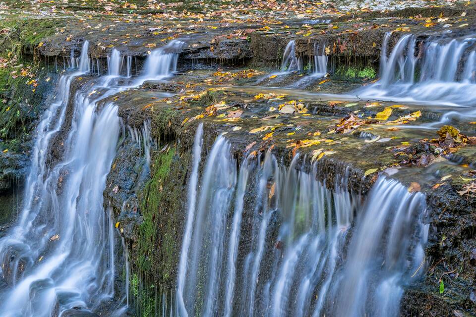 Uma cascata em vários níveis, com rochas cobertas de musgo e folhas de outono.