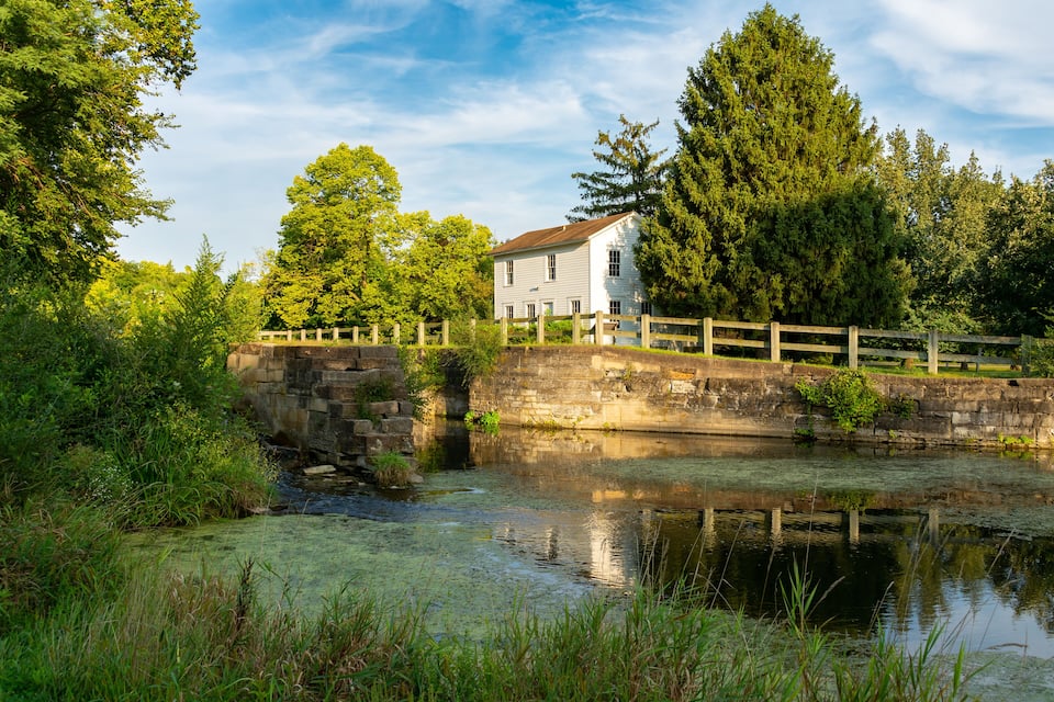 Uma casa histórica branca com uma ponte de pedra sobre um rio calmo, rodeada por vegetação exuberante e árvores altas.