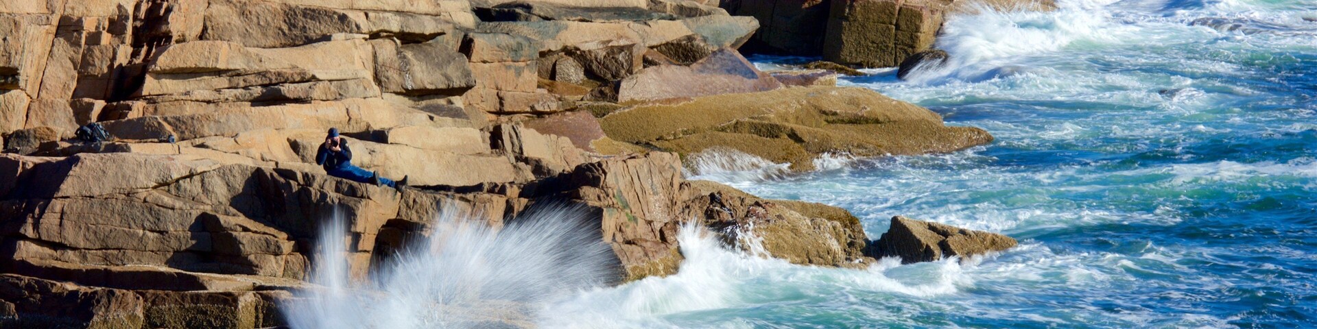 Uma costa rochosa com ondas quebrando nas pedras e uma pessoa fotografando.