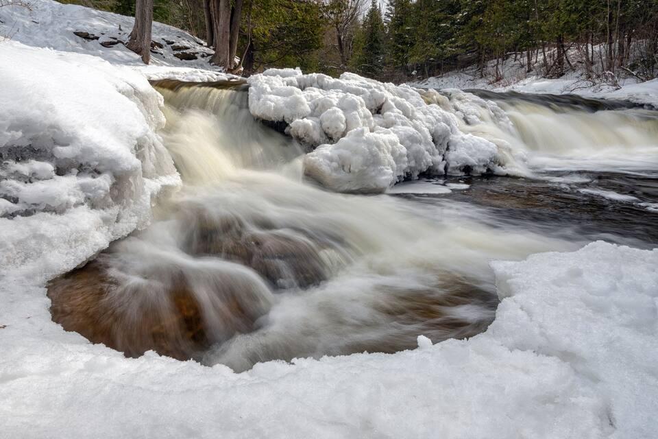 Uma cascata congelada com montes de neve e árvores ao fundo.