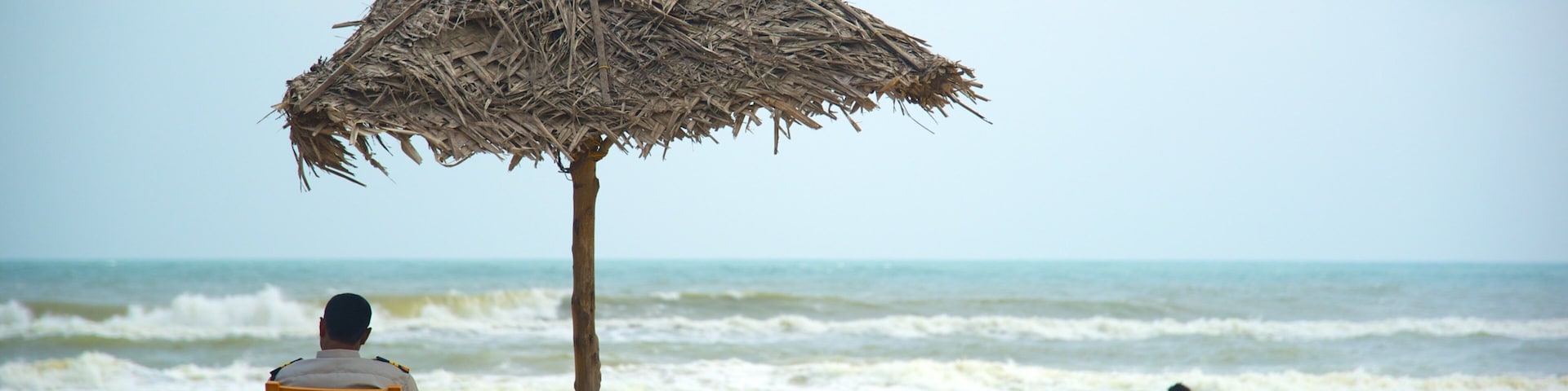 Uma praia com um guarda-sol de palha, um boi de praia e uma pessoa sentada em uma cadeira.