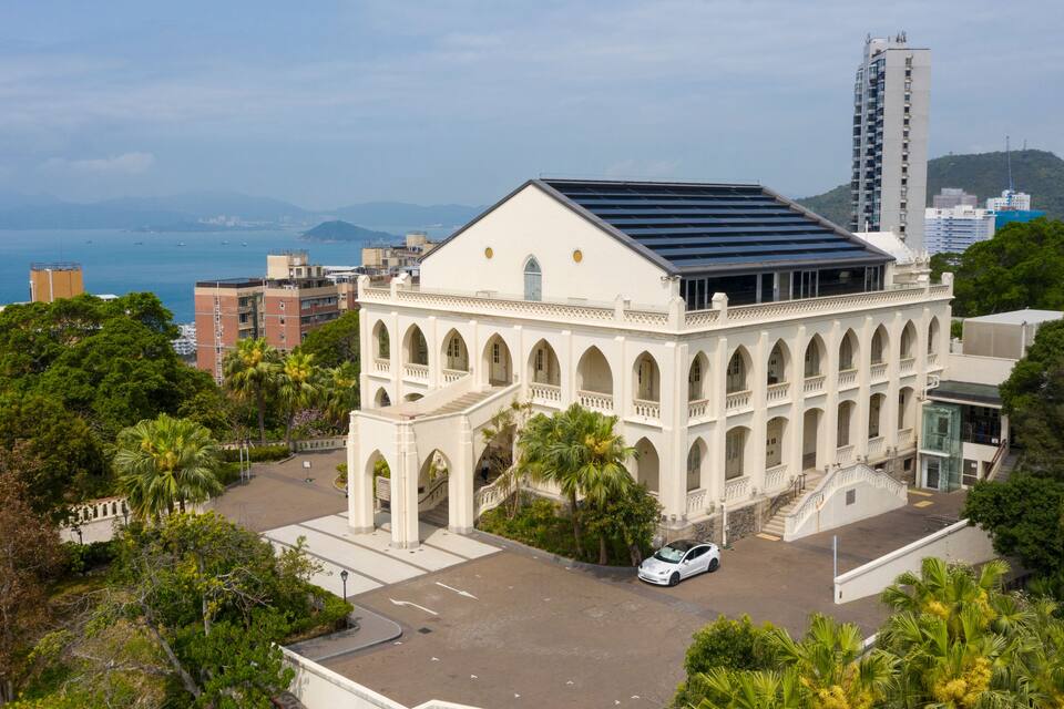 Edifício branco com janelas em arco e telhado com painel solar, cercado por vegetação e com vista para um corpo d'água.