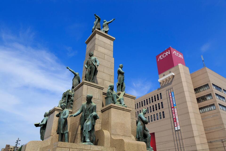 Uma estátua de bronze de figuras históricas em um pedestal, com um edifício moderno e uma placa vermelha ao fundo.