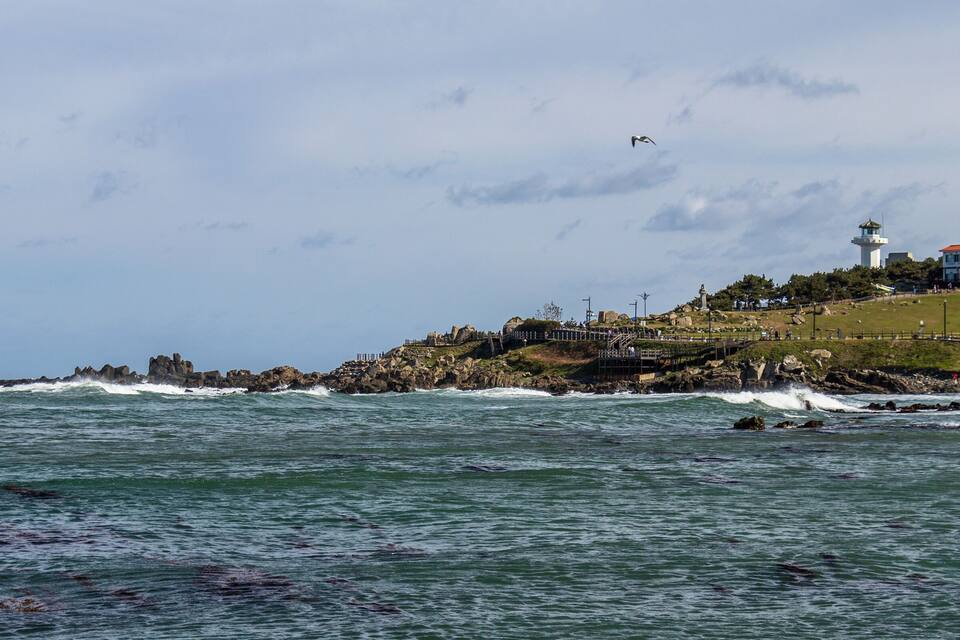 Paisagem costeira com ondas quebrando nas rochas, um farol e uma colina gramada com edifícios.