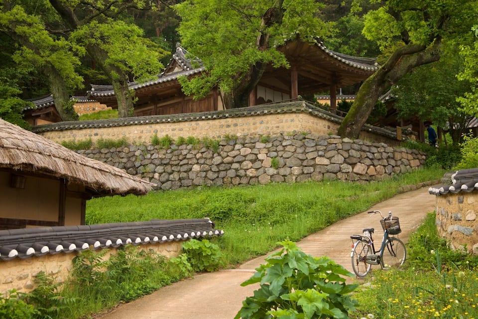 Um jardim coreano tradicional com muros de pedra, construções de telhado de palha e uma bicicleta.