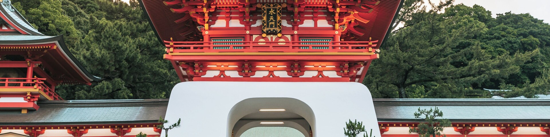 Um templo japonês tradicional com estrutura proeminente em vermelho e branco, rodeado por vegetação exuberante e terreno rochoso.