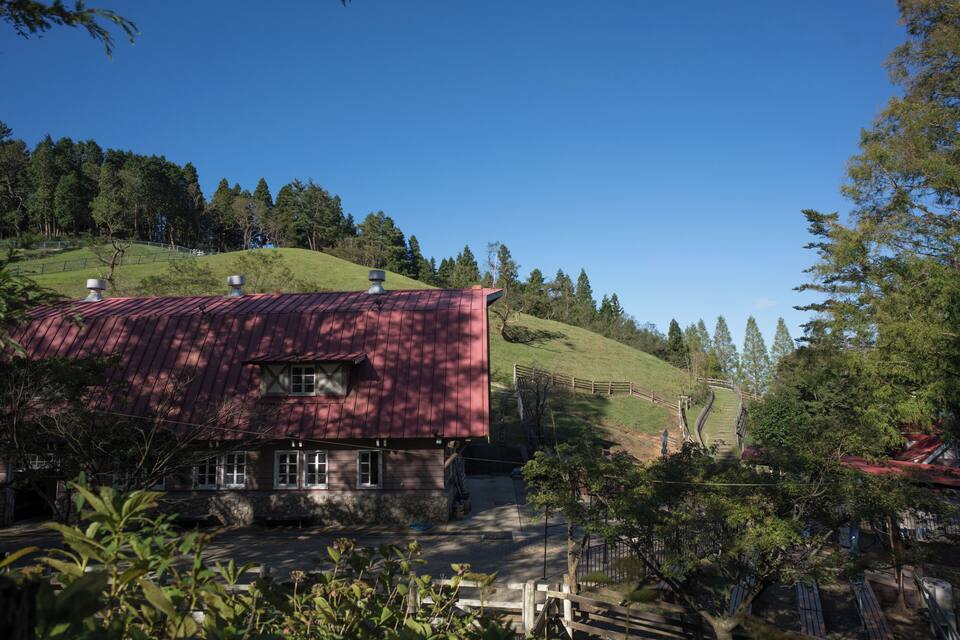 Uma casa com telhado vermelho, parede de pedra e cerca de madeira, numa área arborizada e com declives.