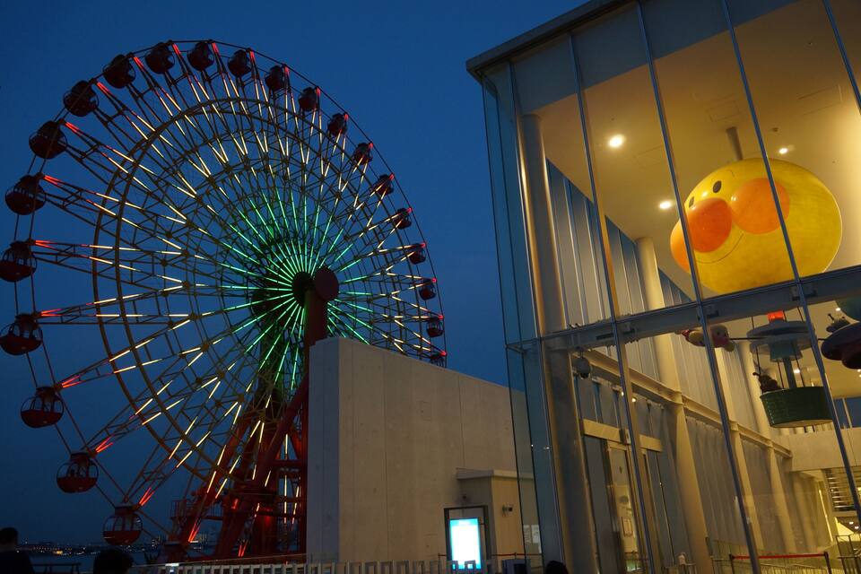 Uma roda gigante iluminada com luzes coloridas ao anoitecer, um edifício com uma grande escultura de pato amarelo e fachada de vidro moderna.