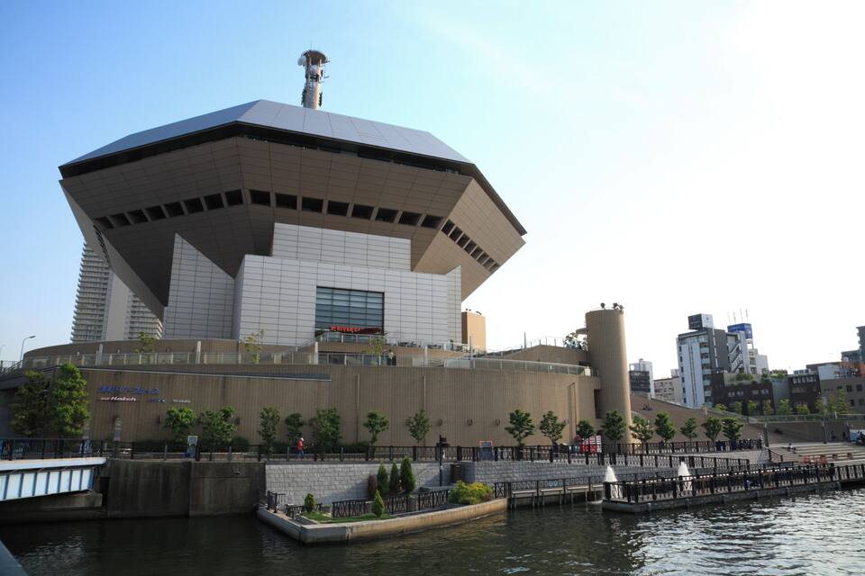 Um edifício moderno com um design geométrico único, localizado à beira-mar, próximo a uma ponte e árvores.