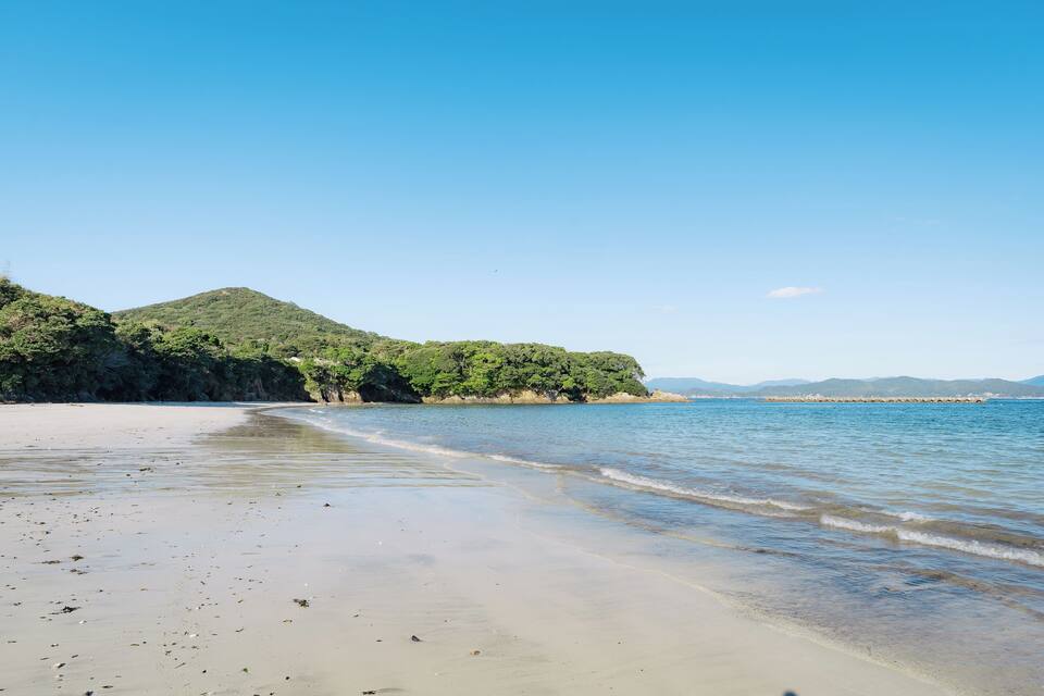 Uma praia com águas azul-claras, areal arenoso e colinas verdes ao fundo.