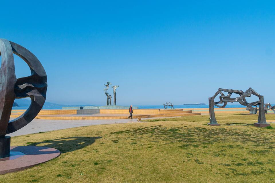 Um parque com esculturas abstratas, uma pessoa passeando com um cachorro, e um céu azul límpido.