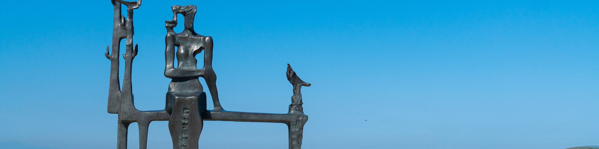 Escultura em bronze de uma mulher e um homem com um pássaro sentados num banco à beira-mar.