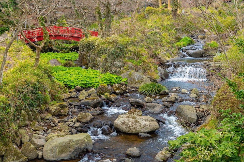 Um riacho com pedras e vegetação, uma ponte vermelha e uma estrutura de madeira.