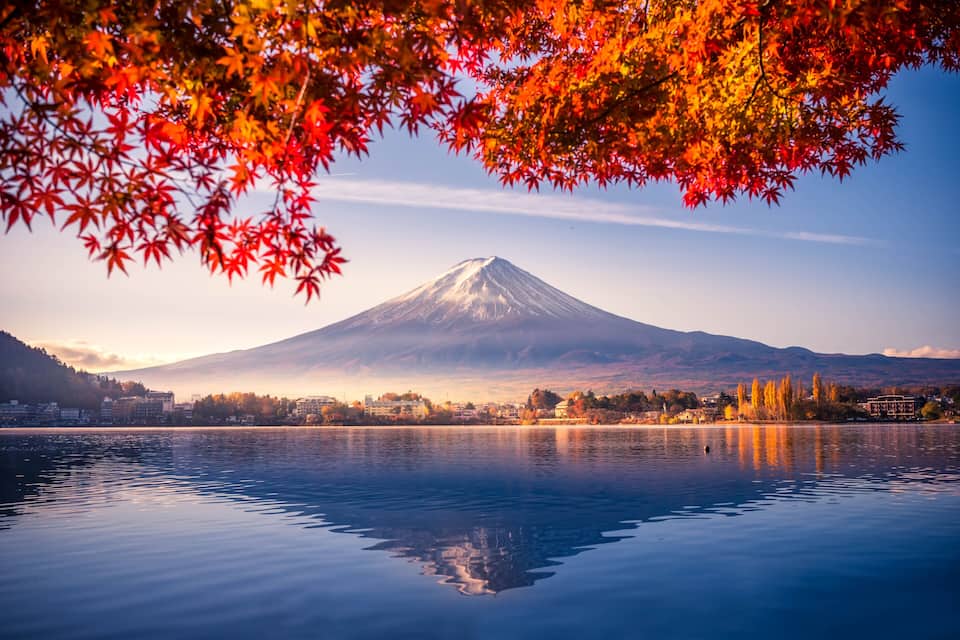 Um lago calmo refletindo uma montanha e folhas de outono em primeiro plano.